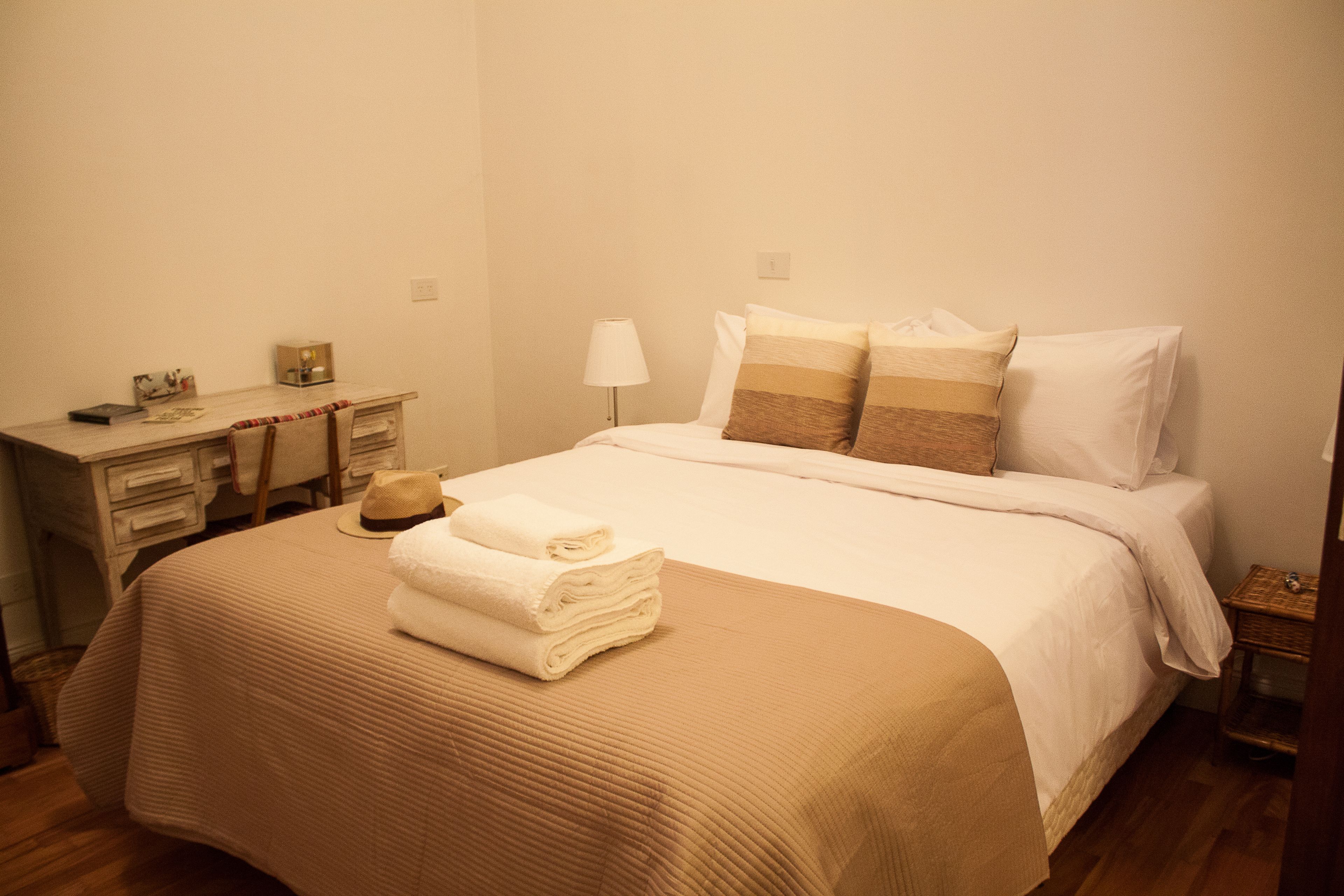Cama bem arrumada com edredom bege e almofadas, mesa de cabeceira pequena com abajur e escrivaninha com chapéu e toalhas dobradas.
