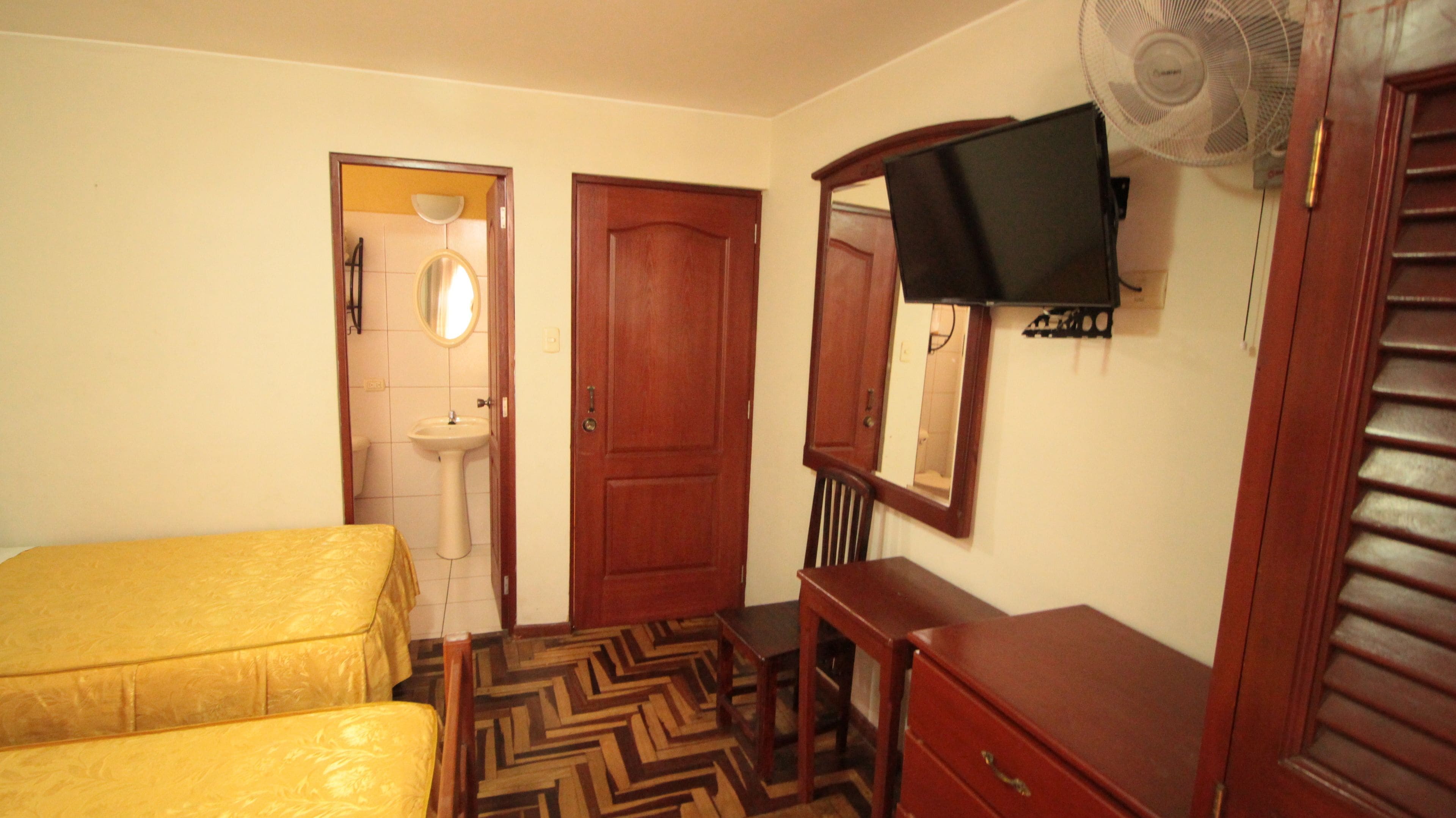 Quarto de hotel pequeno com uma cama, penteadeira de madeira, televisão na parede e banheiro com pia e espelho.