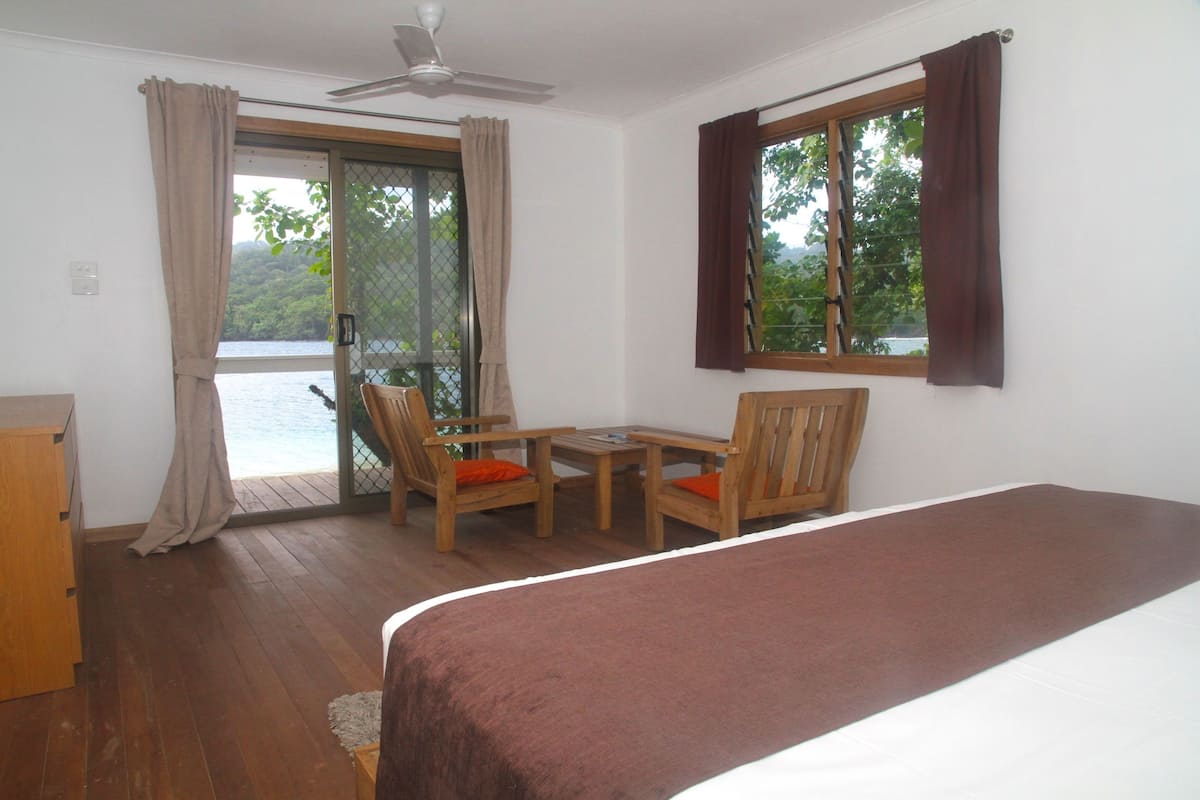 Quarto com cama, uma mesa de madeira, duas cadeiras, janela com cortinas e vista para um corpo de água.