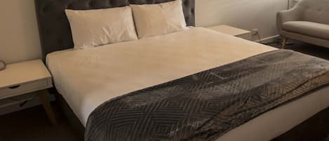 Cama bem arrumada com cabeceira estofada, duas mesas de cabeceira, uma cadeira e um abajur.