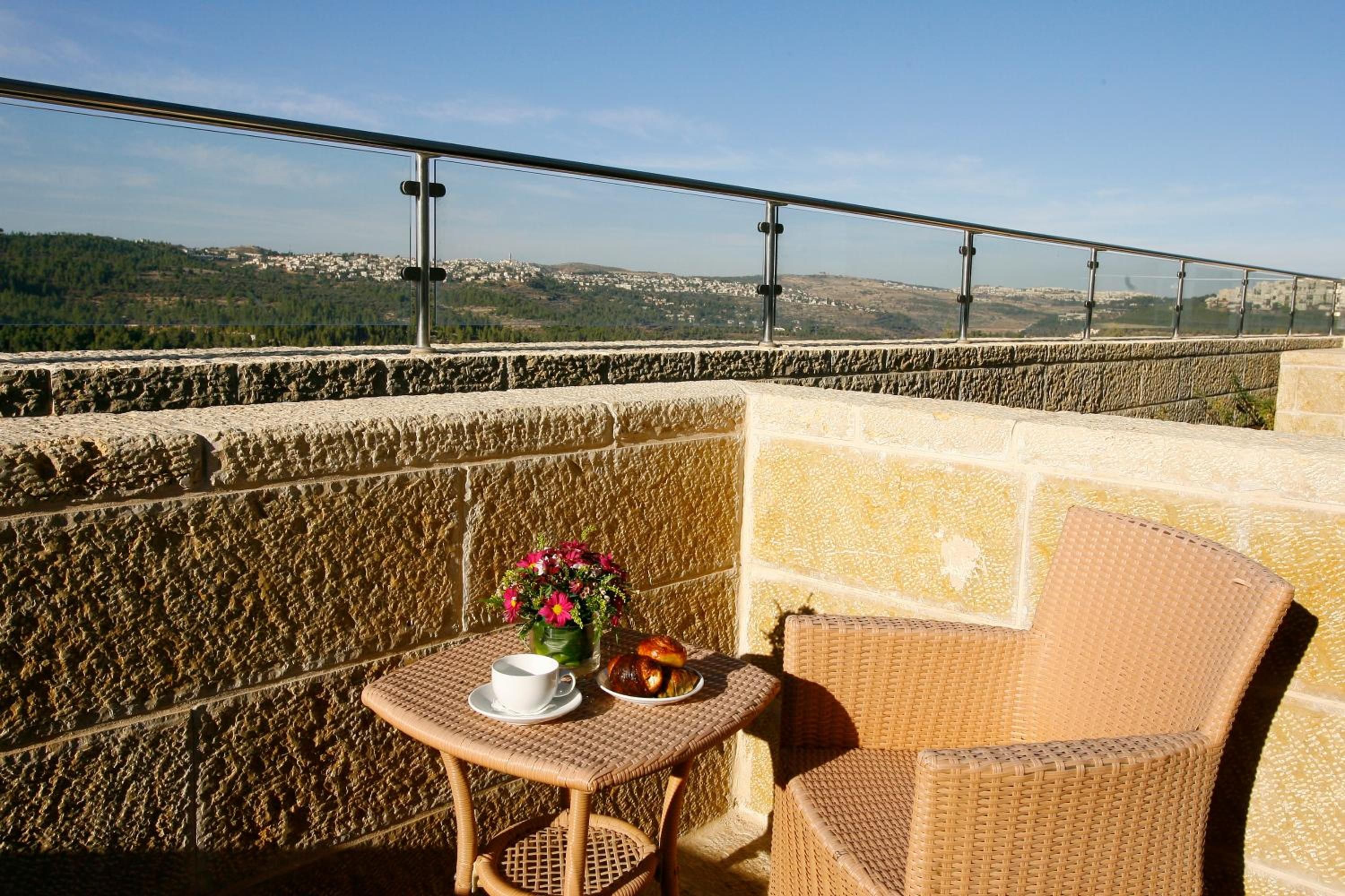 Varanda com paredes de pedra, uma cadeira de vime, uma pequena mesa com uma xícara e flores, e vista para uma cidade ao longe.