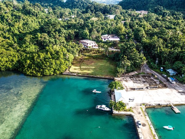 Vista aérea de uma área costeira com um porto de recreio, barcos e alguns edifícios, tudo circundado por uma floresta densa.