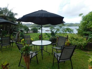 Uma área de estar externa com uma mesa redonda e cadeiras, um grande guarda-sol e vista para um lago com barcos.