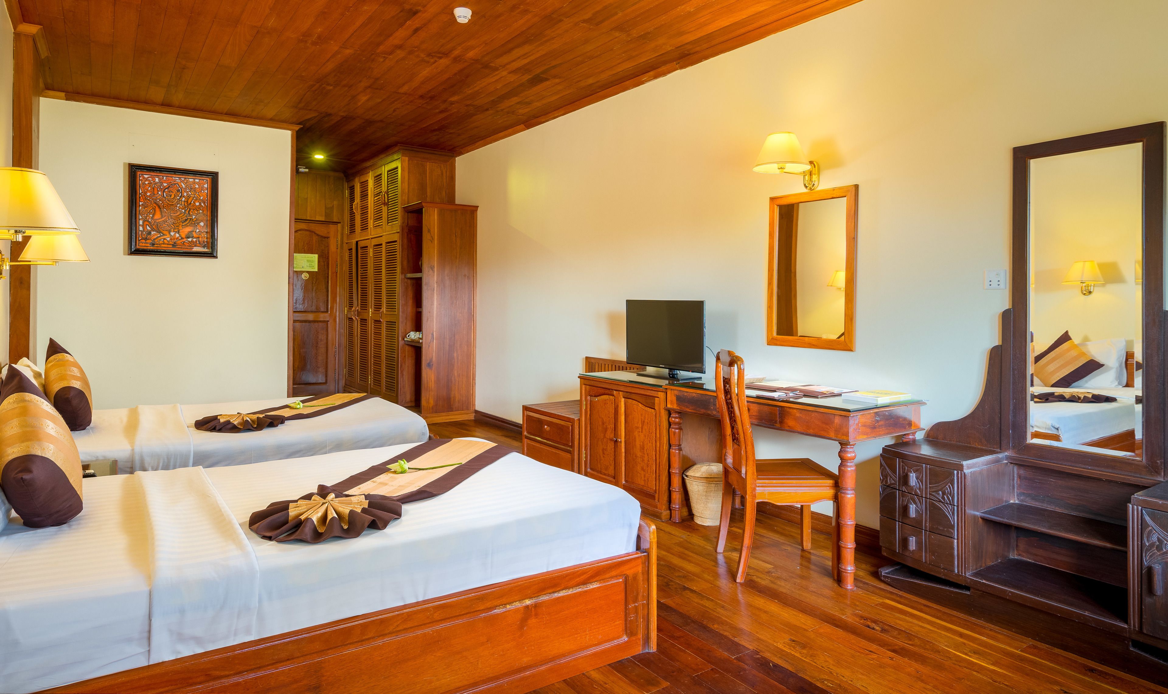 Quarto de hotel com duas camas, uma escrivaninha, uma cadeira, uma televisão e um espelho.