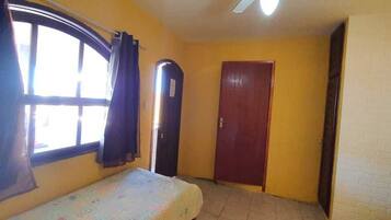 Um quarto com cama, janela com cortinas, porta e parede de tijolos brancos.