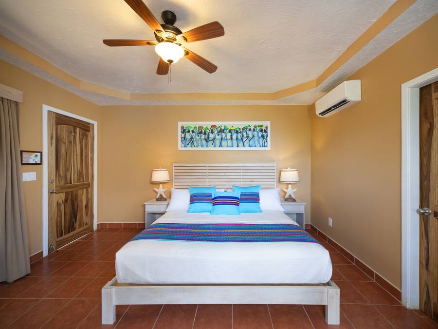 Um quarto com uma cama, duas mesas de cabeceira com abajures, um ventilador de teto, um ar condicionado fixado na parede e uma porta decorada com estrelas.