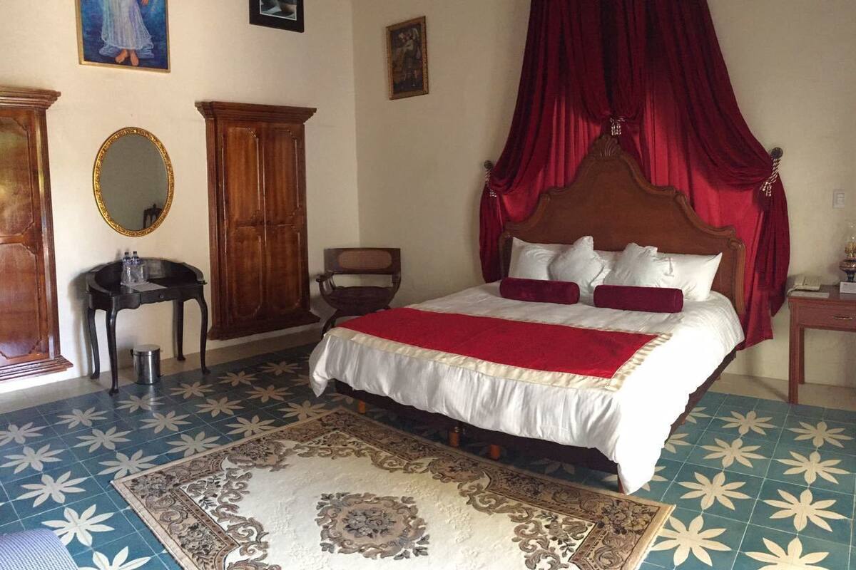 Quarto com uma cama grande, um dossel vermelho, cabeceira de madeira, mesa de cabeceira, uma cadeira, um espelho e um tapete estampado.