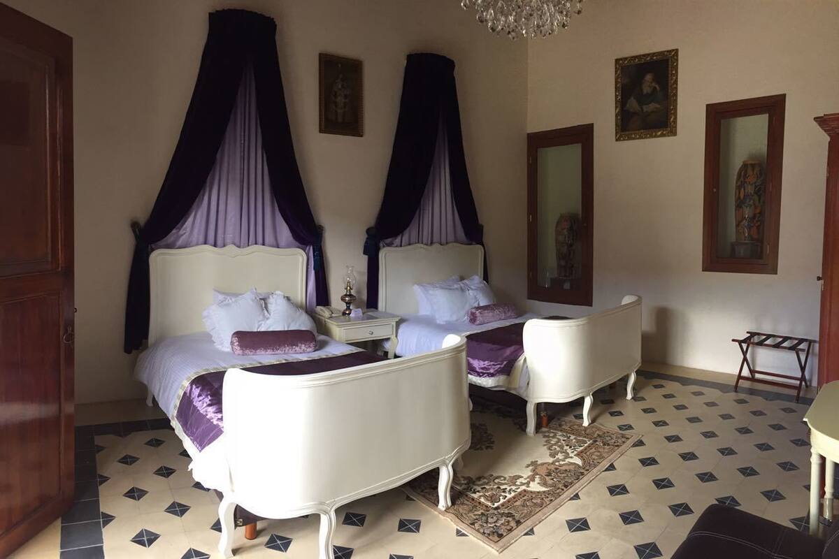 Quarto com duas camas, mesa de cabeceira, cadeira, mesa e lustre.