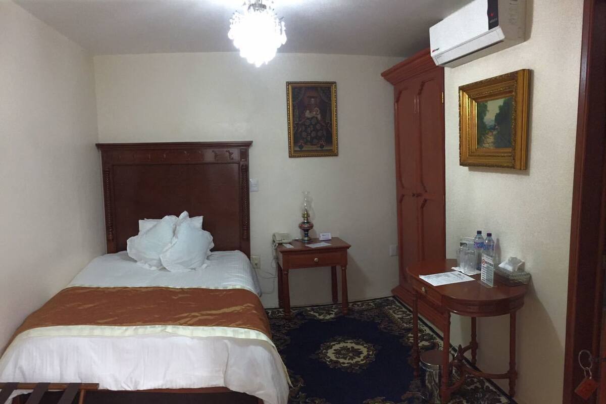 Quarto de hotel com cama, mesa de cabeceira, pequena mesa, cadeira, tapete e guarda-roupa.