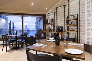 Uma área de refeições com uma mesa de madeira posta para dois, uma garrafa de vinho e uma taça. Cadeiras rodeiam a mesa, e há uma estante com garrafas e taças na parede.