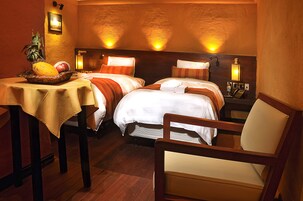 Quarto de hotel com duas camas, uma mesa de jantar com frutas e uma cadeira.