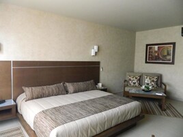 Quarto de hotel com uma cama grande, uma mesa de cabeceira com abajur, uma cadeira e uma pequena mesa com um vaso.