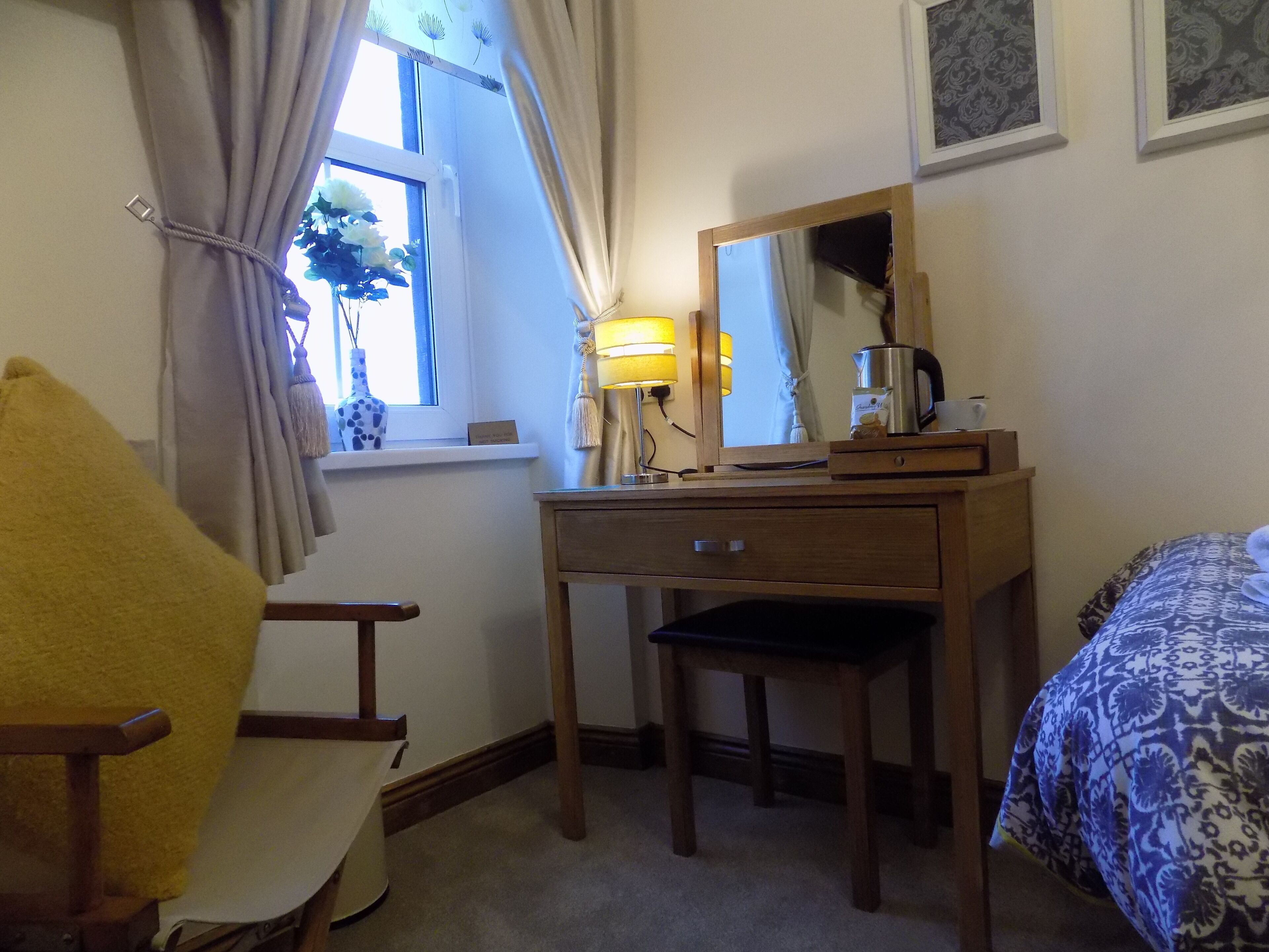 Quarto de hotel com escrivaninha de madeira, uma cadeira, cama com roupa de cama estampada, janela com cortinas e espelho fixado na parede.
