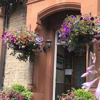 Entrada de um edifício com cestos de flores pendurados e uma porta com uma placa.