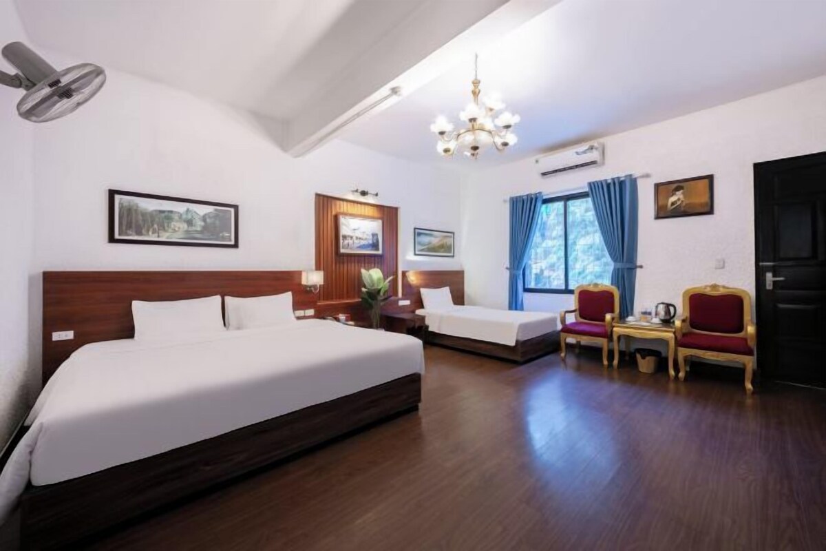 Quarto de hotel com uma cama grande, duas camas menores, uma escrivaninha, duas cadeiras, um candelabro e quadros nas paredes.