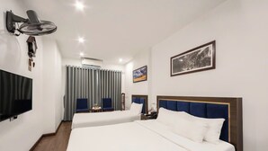 Quarto de hotel com duas camas, uma TV de tela plana, um ventilador e uma janela com cortinas, além de um quadro pendurado na parede.