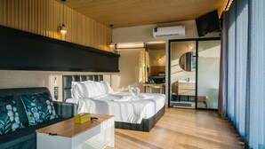 Quarto de hotel moderno com teto de madeira, cama com lençóis brancos, mesa de cabeceira de madeira, sofá com almofadas florais e banheiro visível por uma porta de vidro.