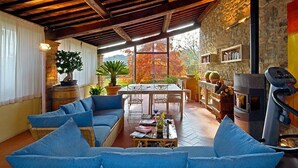 Uma sala de estar acolhedora com parede de pedra, vigas de madeira no teto, lareira e um espaço de jantar com mesa e cadeiras.
