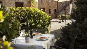 Mesa de jantar ao ar livre com pratos de comida, uma garrafa de vinho e um jarro, cercada por vegetação e um edifício de pedra ao fundo.