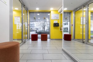 Uma área de recepção com parede amarela, portas de vidro e piso ladrilhado. Há três pufes vermelhos e uma cadeira marrom.