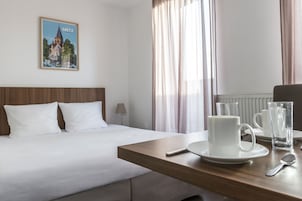 Quarto de hotel com cama, cabeceira de madeira, mesa com caneca branca e copo, e pôster de uma igreja na parede.