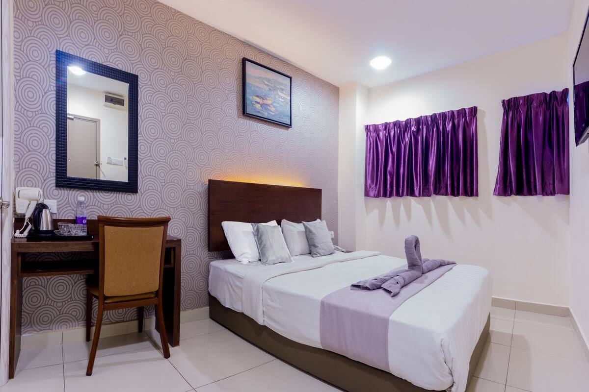 Quarto de hotel com cama, escrivaninha, cadeira, espelho e cortinas roxas.