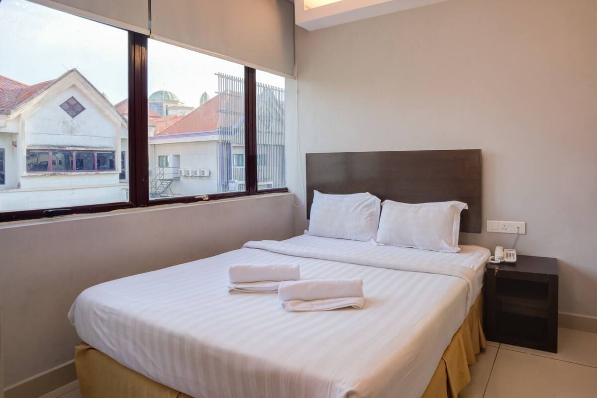 Quarto de hotel com uma cama grande, uma mesinha de cabeceira com telefone e uma janela com vista para prédios.