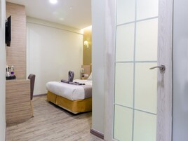 Quarto de hotel com cama, mesa de cabeceira, abajur e chão de madeira.