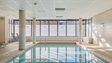Piscina interna com paredes de vidro transparente, piso ladrilhado e teto com iluminação embutida.