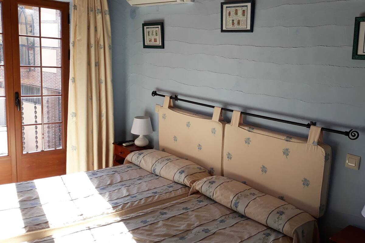 Quarto com cama, mesa de cabeceira, luminária, janela com cortinas e quadros na parede.