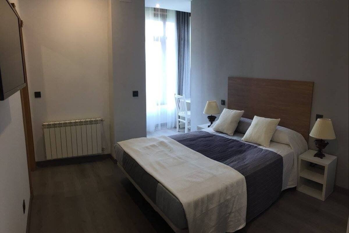 Quarto com uma cama grande, duas mesas de cabeceira com abajures, um radiador e uma janela com cortinas.