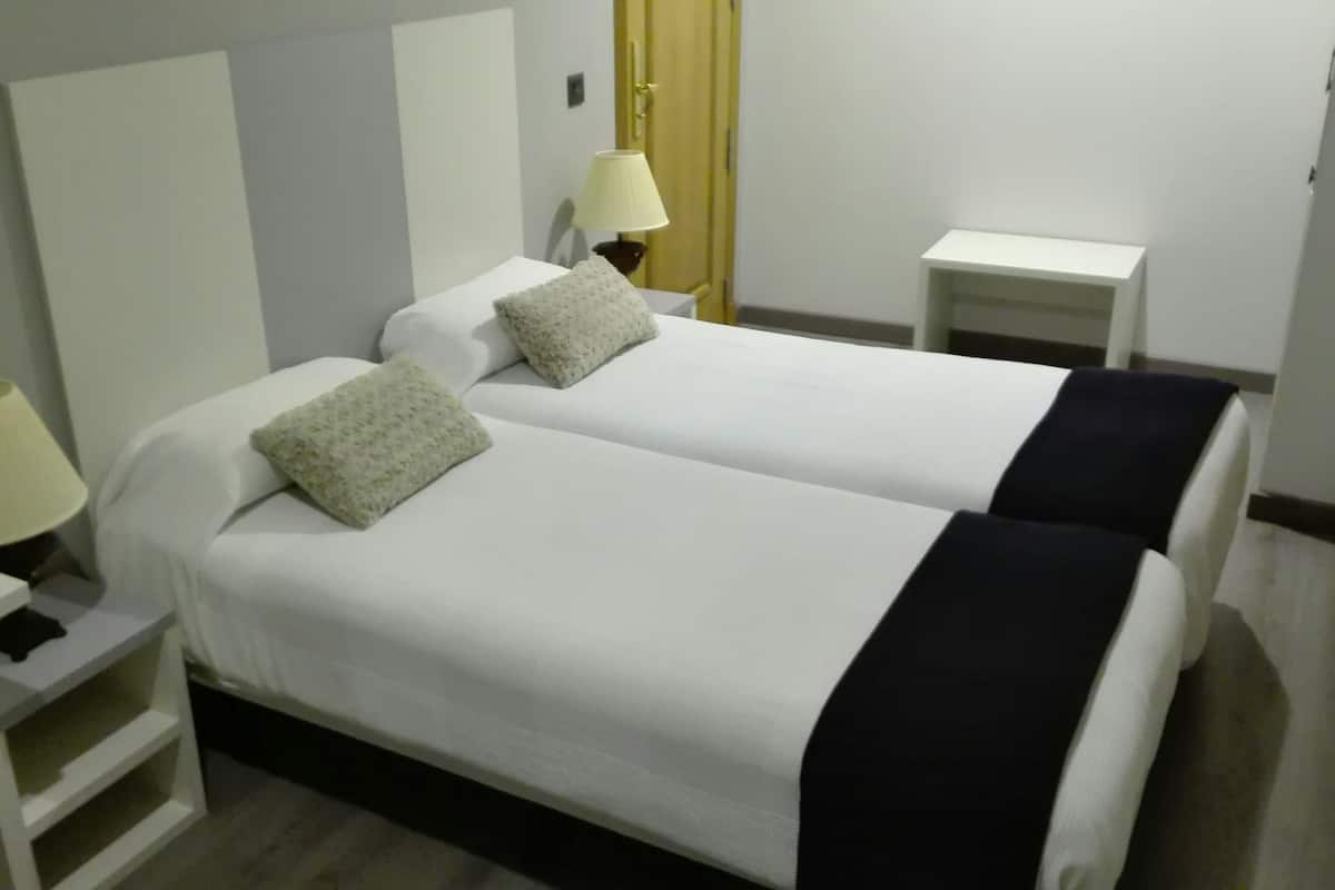 Quarto de hotel com duas camas, uma mesinha de cabeceira, um abajur e uma porta.