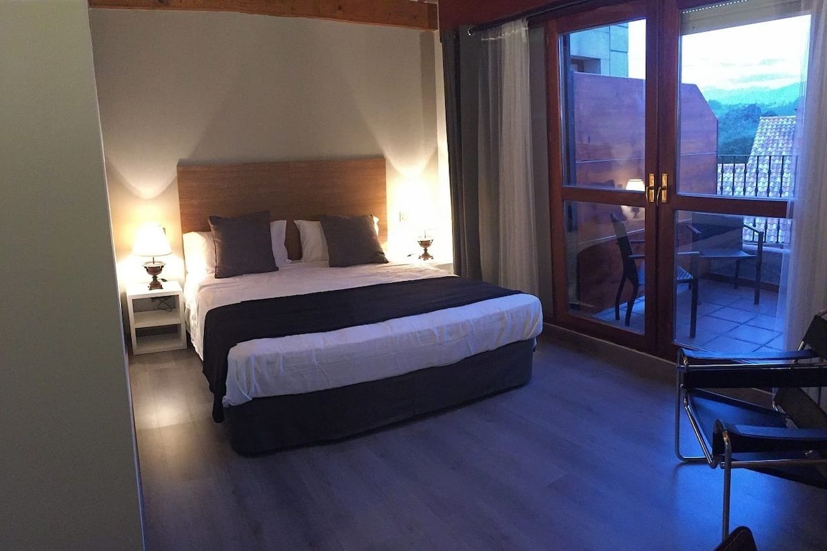 Quarto com cama grande, mesa de cabeceira, abajur e vista para área externa por uma porta de vidro.