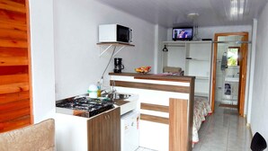 Cozinha compacta com fogão a gás, micro-ondas e pequena geladeira. Na área de estar, há um sofá e uma cadeira, e uma televisão fixada na parede.