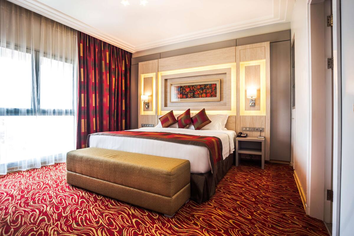 Quarto de hotel com uma cama grande, uma mesinha de cabeceira, um banco, uma janela com cortinas leves e um tapete estampado.