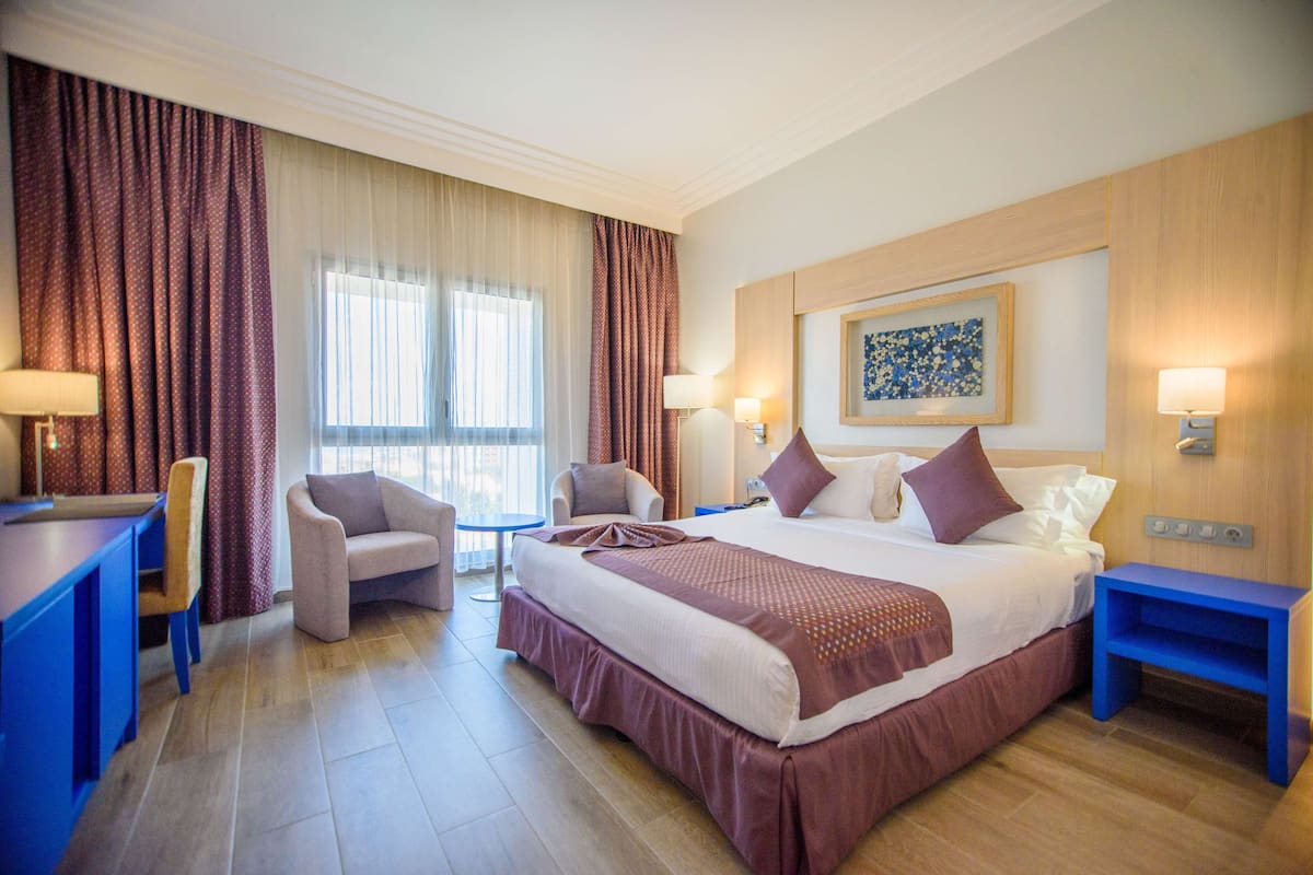 Quarto de hotel com cama grande, duas mesas de cabeceira, escrivaninha, cadeira e janela com cortinas.