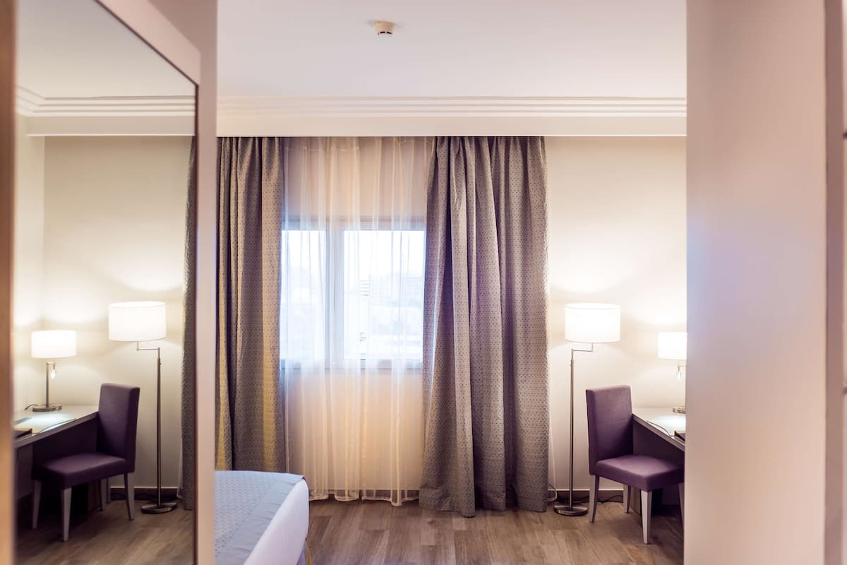 Quarto de hotel com cama, duas cadeiras, escrivaninha, luminária e uma janela grande com cortinas.