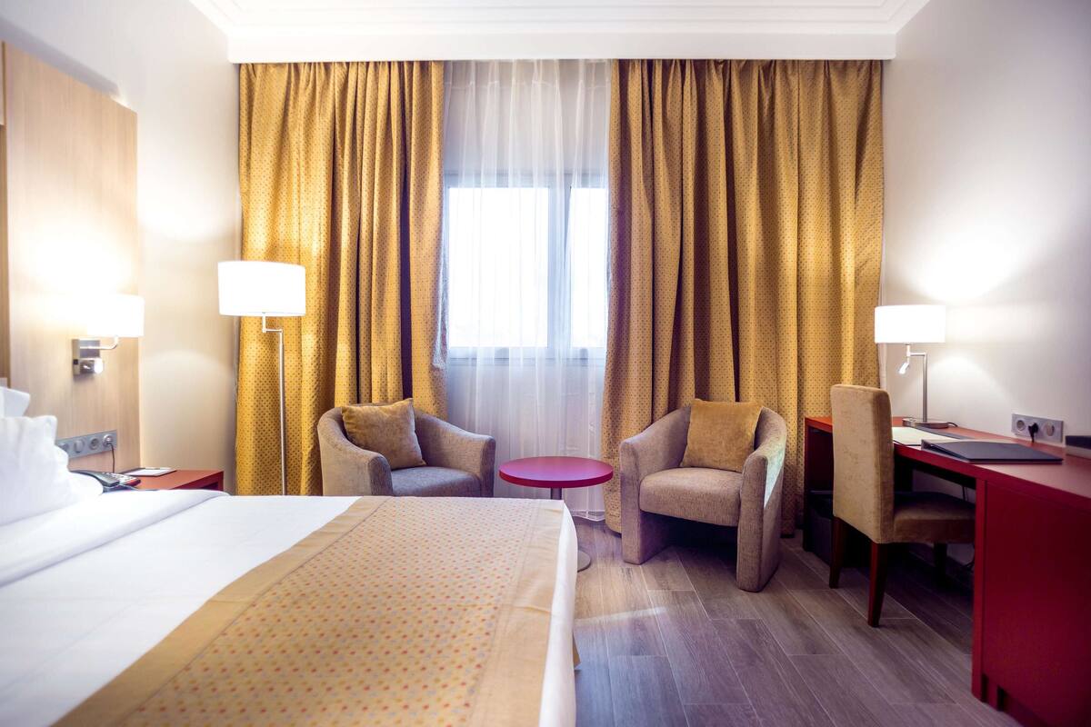 Quarto de hotel com uma cama, duas poltronas, uma mesinha redonda, uma escrivaninha e uma cadeira.
