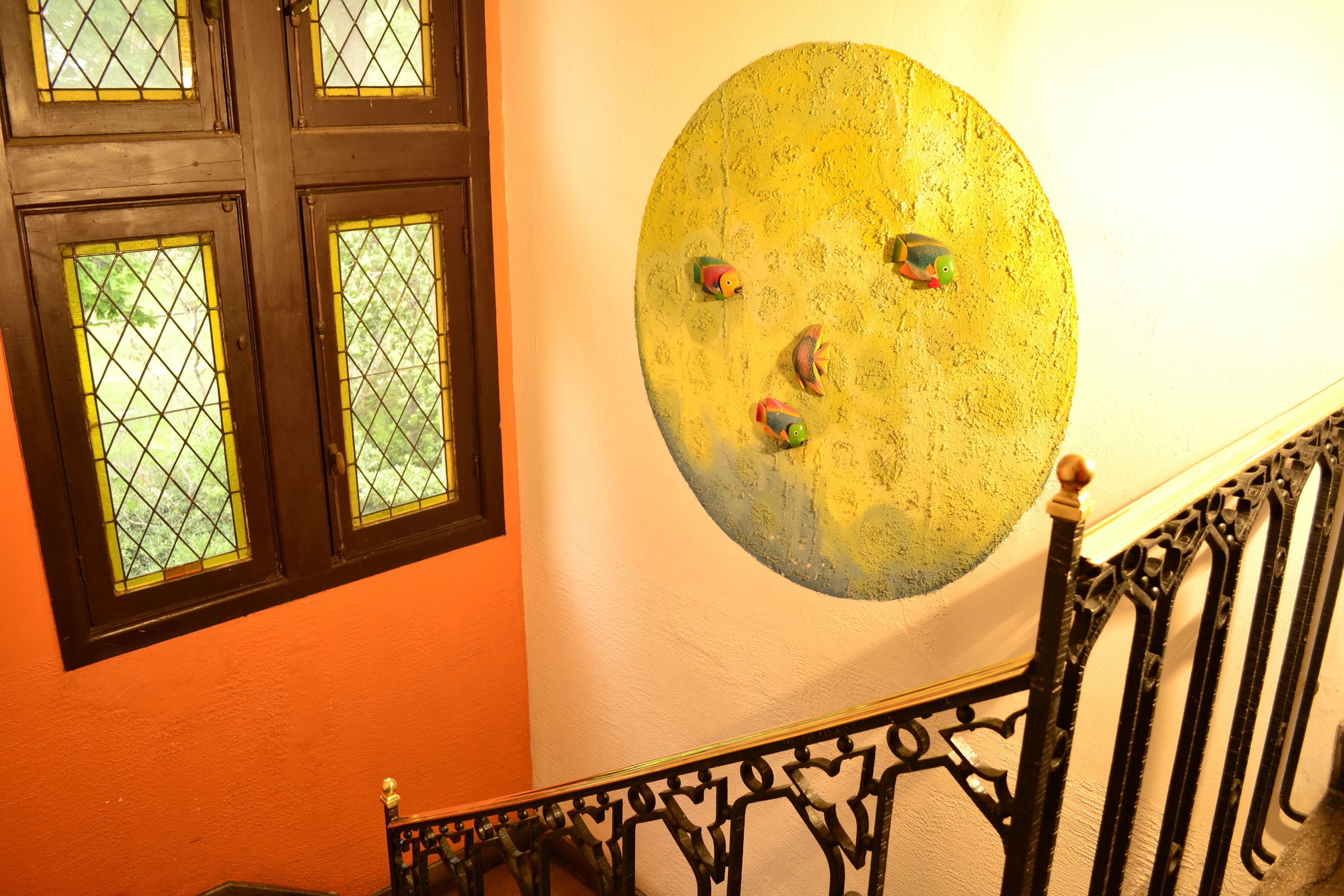 Uma escadaria com grades pretas ricamente decoradas e uma parede com uma obra de arte circular amarela retratando peixes coloridos.