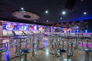 Um bar espaçoso com vários bancos altos e mesas, teto iluminado e um mural vibrante com tema espacial na parede.
