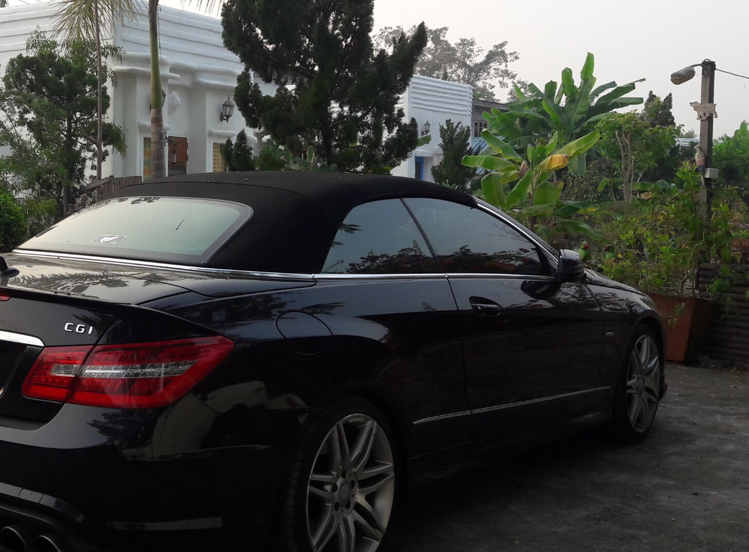 Um carro conversível preto estacionado numa entrada de garagem, com um edifício branco e vegetação exuberante ao fundo.