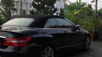Um carro conversível preto estacionado numa entrada de garagem, com um edifício branco e vegetação exuberante ao fundo.