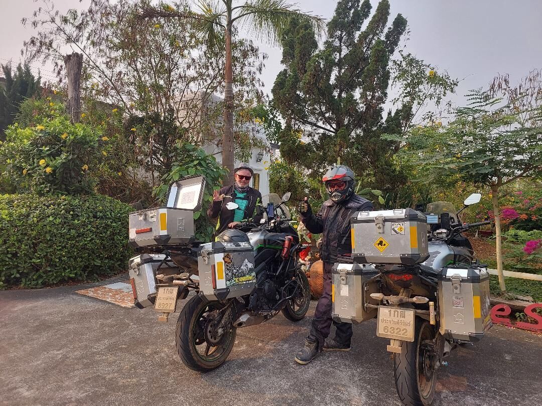 Duas motocicletas estacionadas ao ar livre, com caixas de armazenamento, rodeadas por vegetação e árvores.