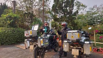 Duas motocicletas estacionadas ao ar livre, com caixas de armazenamento, rodeadas por vegetação e árvores.