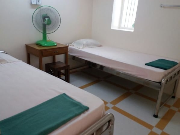 Quarto de hospital com duas camas, um ventilador verde, uma janela e chão de azulejos.