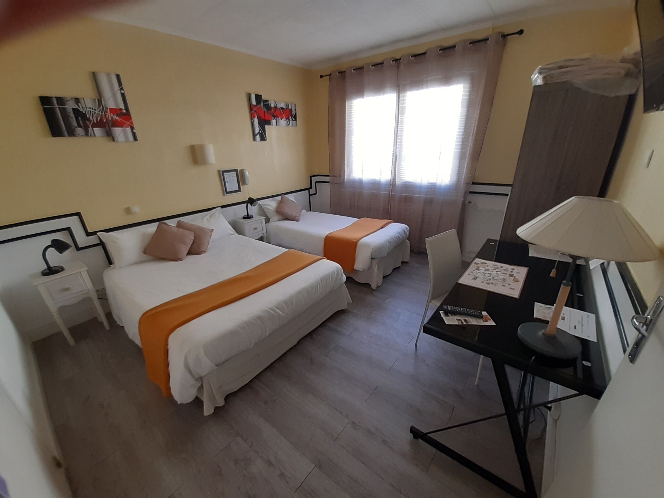 Quarto de hotel com duas camas, uma escrivaninha, uma cadeira, um abajur e uma janela com cortinas.