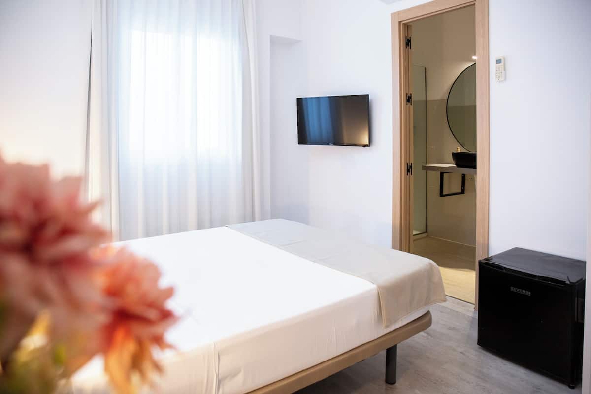 Quarto de hotel com uma cama, uma televisão na parede, um espelho e uma mala preta no chão.