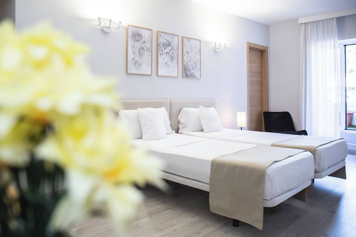 Quarto de hotel com duas camas, porta de madeira, uma cadeira e quadros na parede.