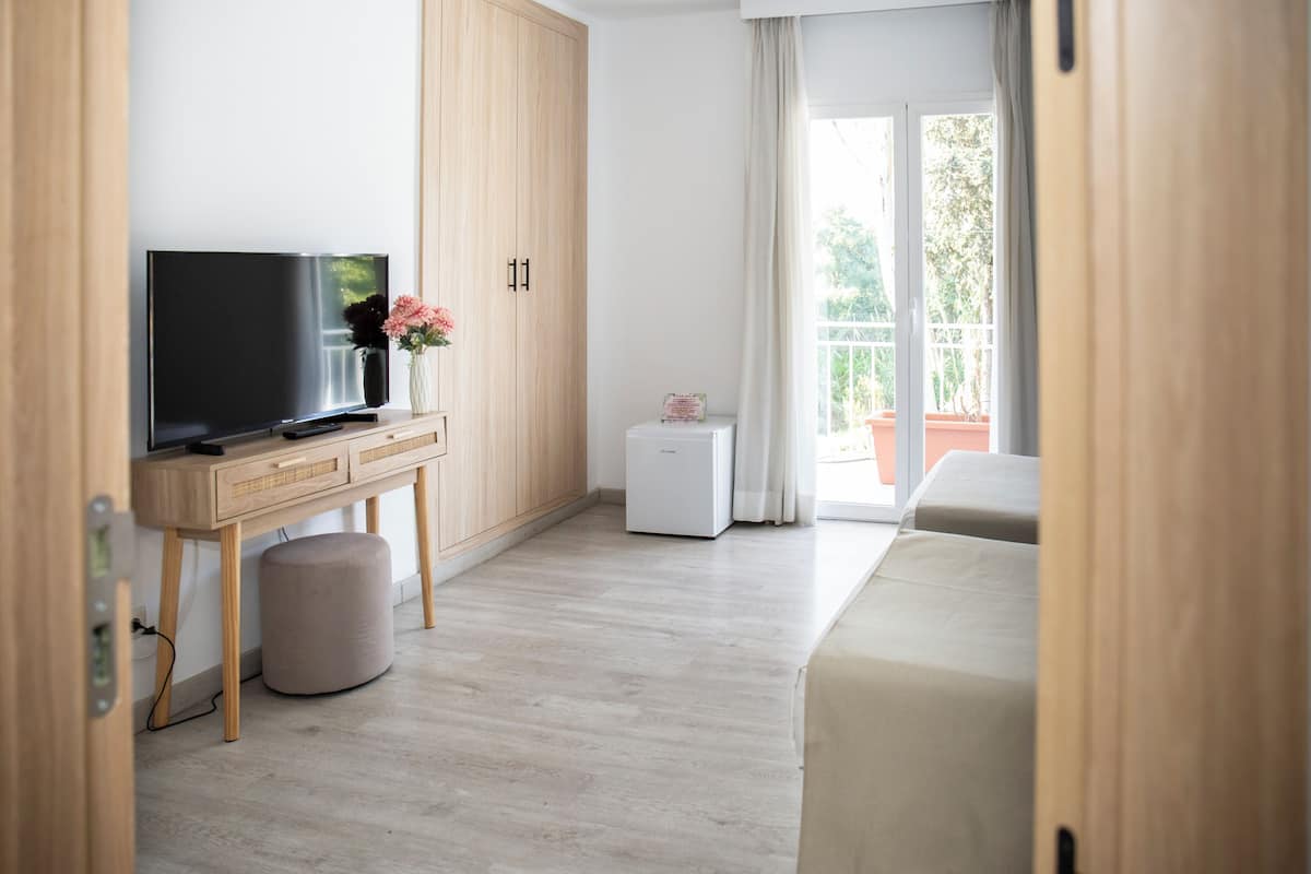 Quarto de hotel moderno com televisão de tela plana, uma mesinha com um vaso de flores, um mini-refrigerador, uma cama e uma varanda com vista para o verde.