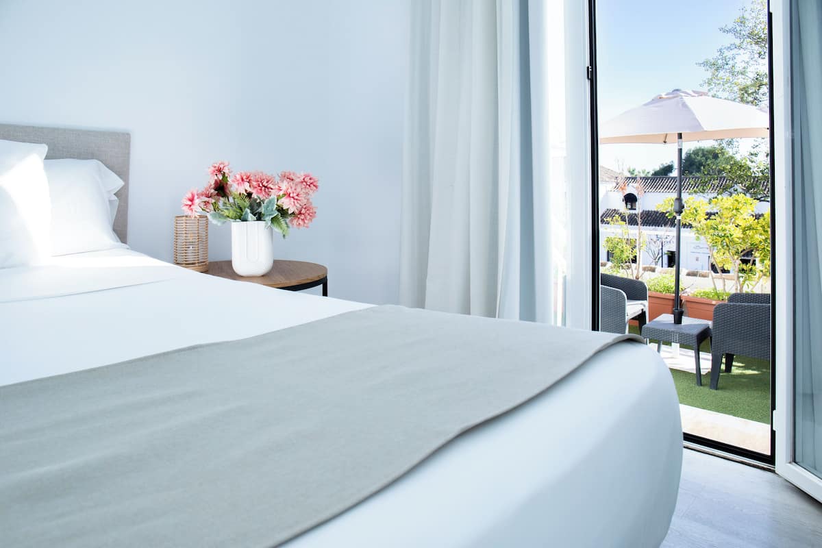 Quarto de hotel com uma cama grande, uma mesinha com um vaso de flores e vista para um pátio externo com assentos.